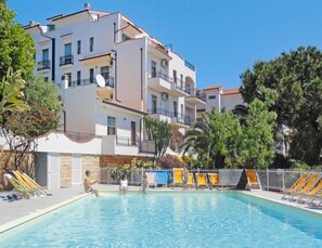 Piscina com espreguiçadeiras, rodeada por um edifício branco com varandas e um céu azul límpido.