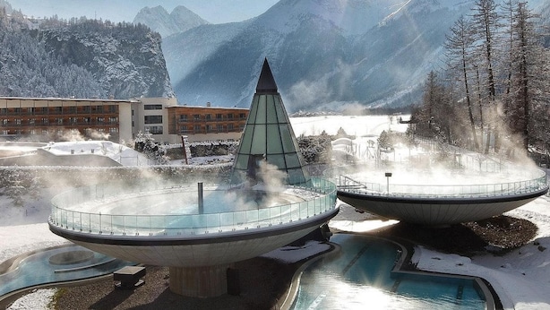 Uma área de spa com uma fonte central, rodeada por montanhas cobertas de neve e um edifício com várias janelas.
