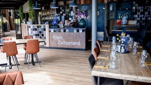 Interior de restaurante com piso de madeira, cadeiras de couro marrom, uma longa mesa de jantar posta com copos e um balcão com a placa 'Klein Schweizland'.