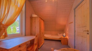 Quarto de cabana de madeira com uma cama de solteiro, uma escrivaninha com gavetas, uma cadeira e um guarda-roupa.