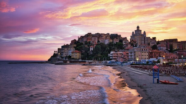 Paisagem urbana costeira ao pôr do sol, com edifícios ao longo da orla e uma praia com guarda-sóis.