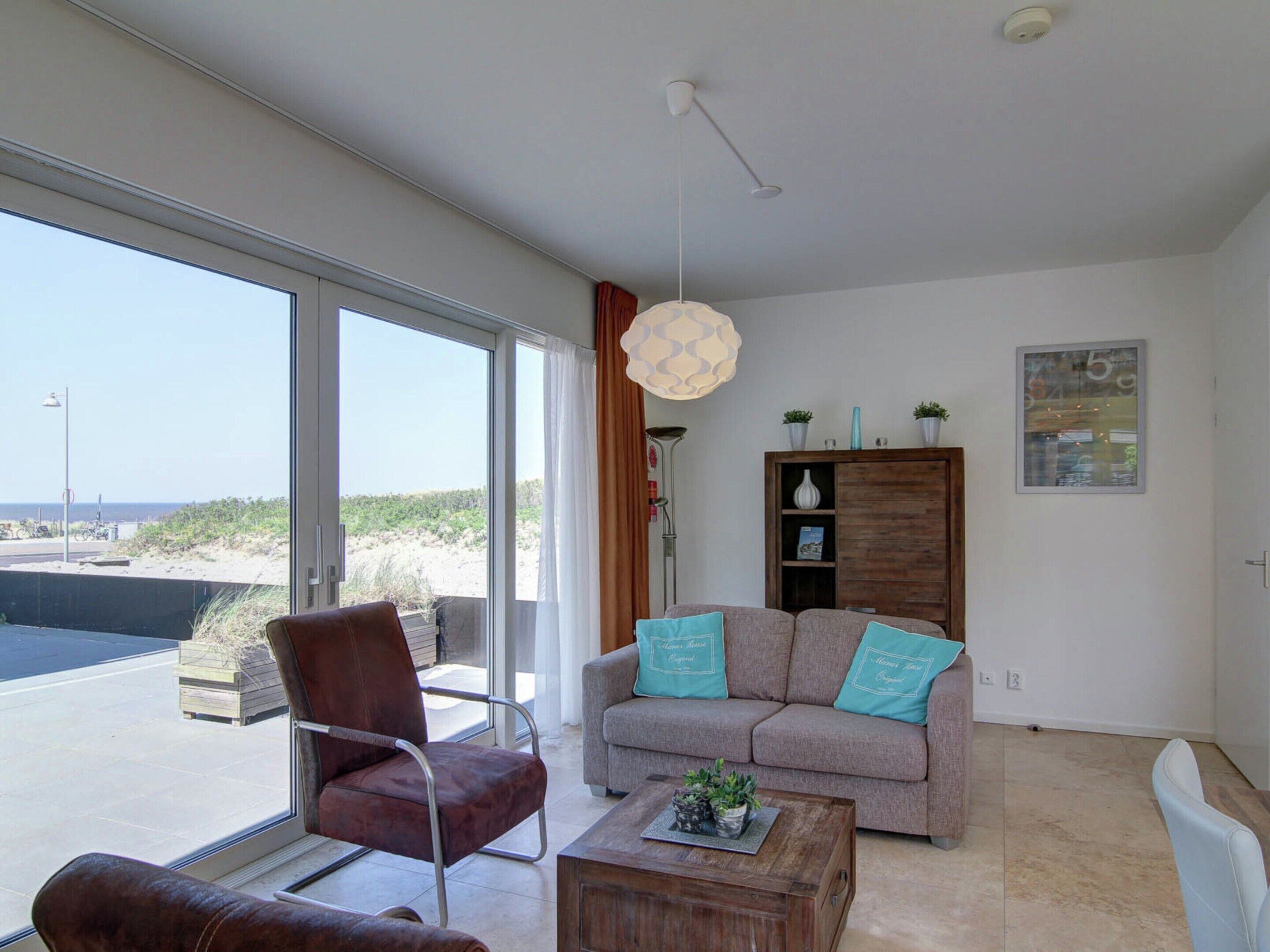 Uma sala de estar moderna com sofá, poltrona e mesa de centro. Há uma janela grande com vista para a praia.
