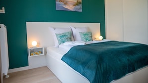 Uma cama bem arrumada com um edredom verde-água, duas almofadas estampadas com folhas e uma mesa de cabeceira com uma luminária.