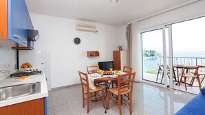 Cozinha compacta com fogão, pia e área de refeições com cadeiras e mesa. Há uma TV e um relógio na parede. O quarto possui uma grande porta de vidro deslizante que dá para uma varanda com mobiliário externo.