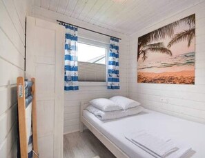 Quarto pequeno com cama de solteiro, janela com cortinas azuis e brancas, painel decorativo com tema praiano e escada de madeira encostada na parede.