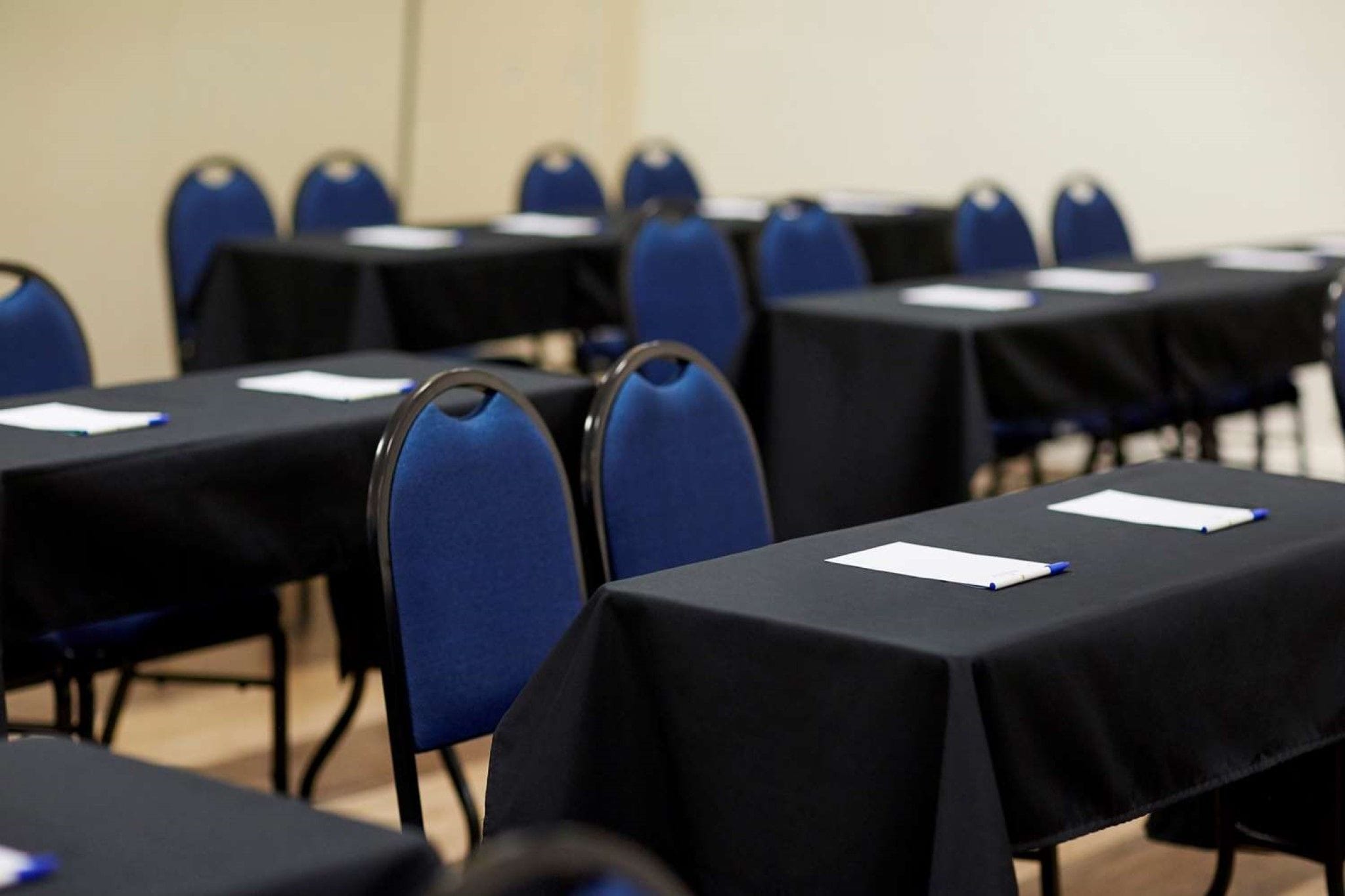 Uma sala de conferências com mesas alinhadas e cadeiras azuis. Cada mesa tem uma toalha preta e papéis sobre ela.