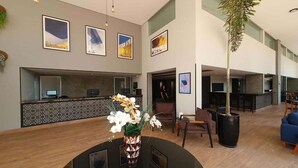 Um saguão de hotel com uma mesa de recepção, obras de arte emolduradas nas paredes, uma grande planta em vaso e uma área de estar com cadeiras e um sofá.