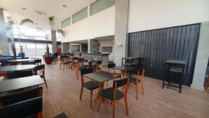 Uma ampla área de refeições interna com várias mesas e cadeiras, uma cozinha e grandes janelas que permitem entrada de luz natural.