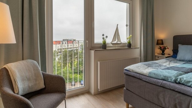 Quarto com cama, uma cadeira, janela com vista para edifícios, radiador e mesa de cabeceira com abajur.