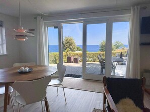 Uma área de refeições com mesa e cadeiras, vista para o mar através de grandes portas de vidro e um lustre.