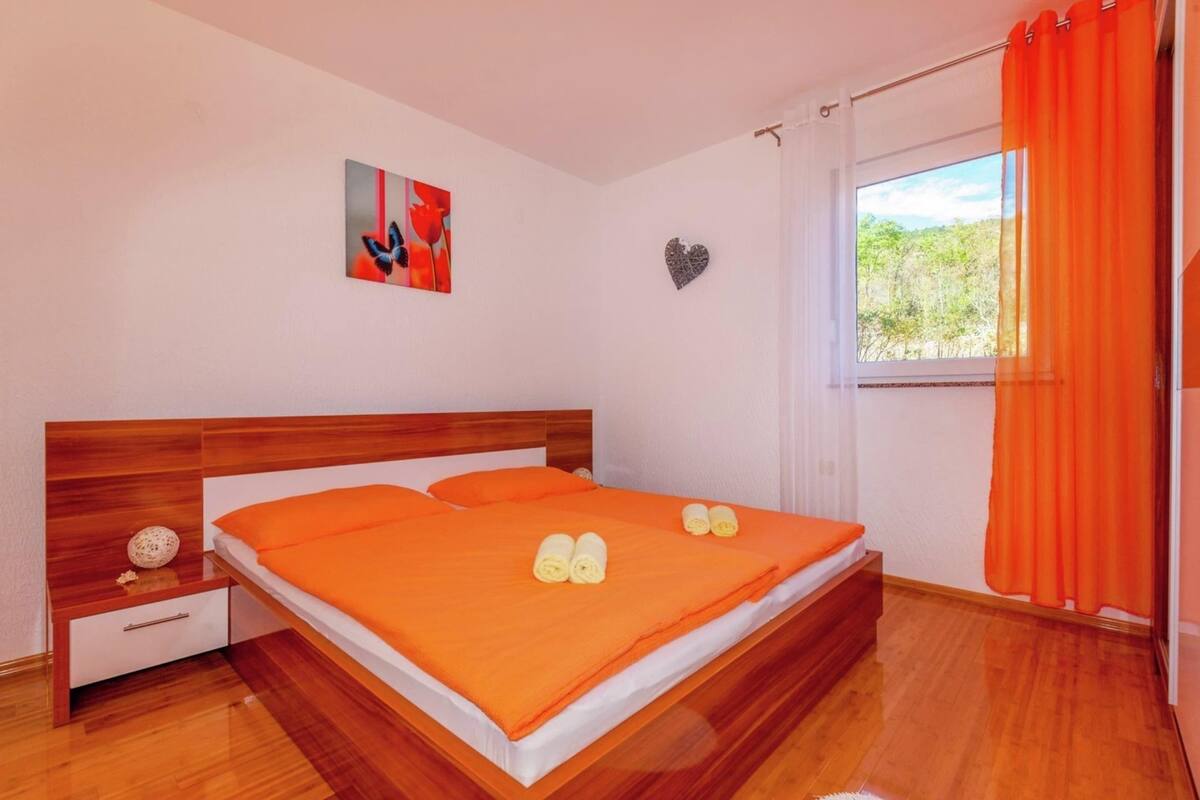 Quarto com cama de madeira, paredes brancas, uma pequena mesa de cabeceira, cortinas laranja na janela e vista para o verde lá fora.