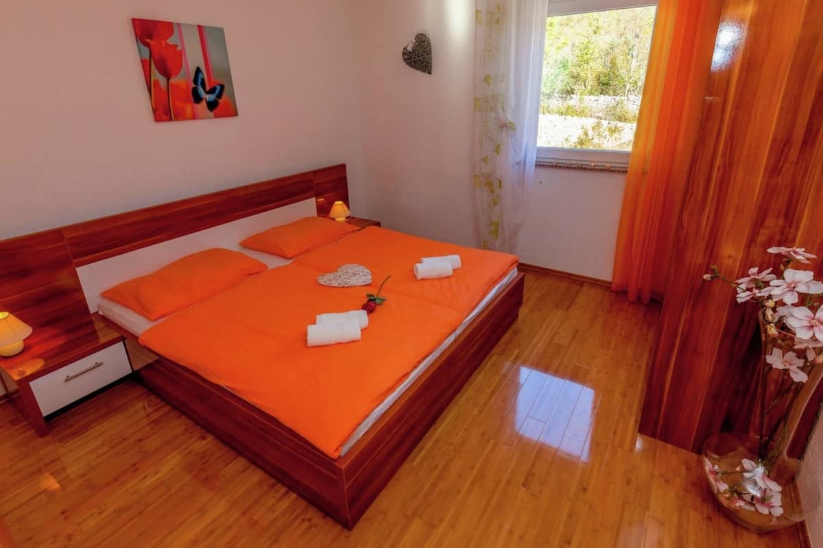 Quarto com cama de madeira, roupa de cama laranja, mesa de cabeceira com abajur, um objeto decorativo na parede e janela com cortinas.