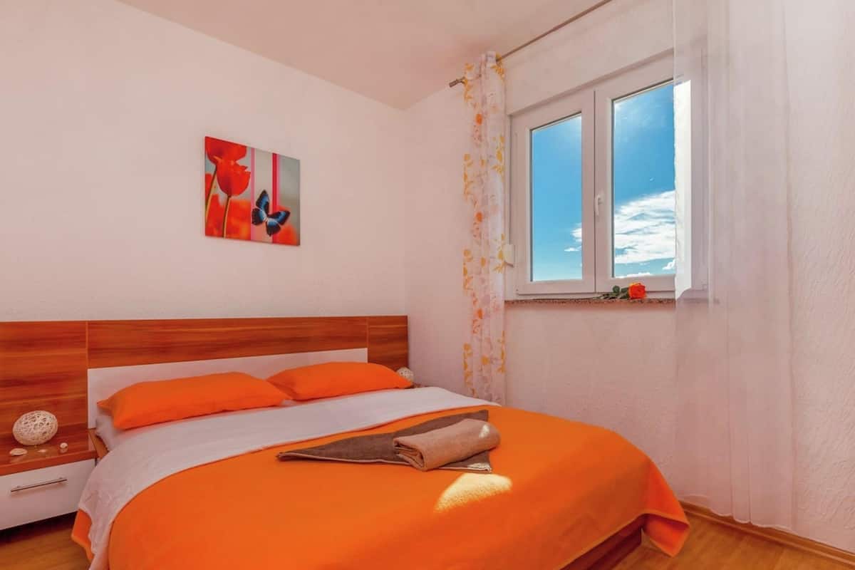 Quarto com cabeceira de madeira, cama com roupa de cama laranja, mesa de cabeceira com objeto decorativo e janela com vista para o céu.