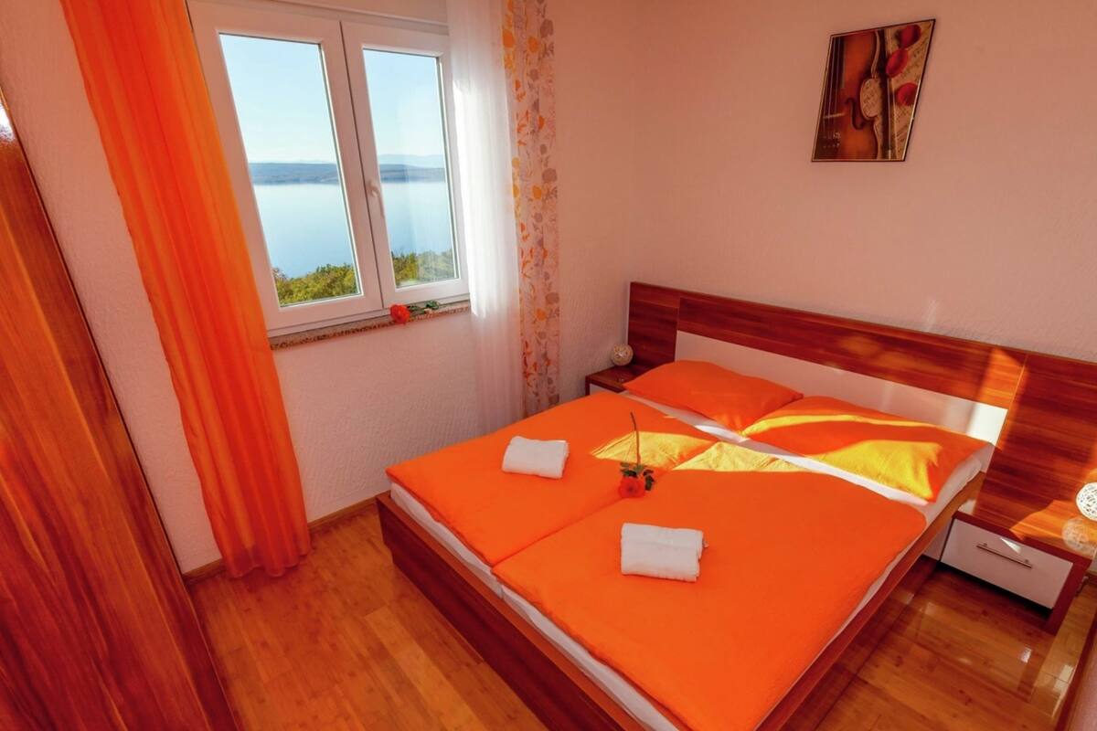 Cama de casal com roupa de cama laranja, cabeceira de madeira, mesa de cabeceira e janela com vista para a água.