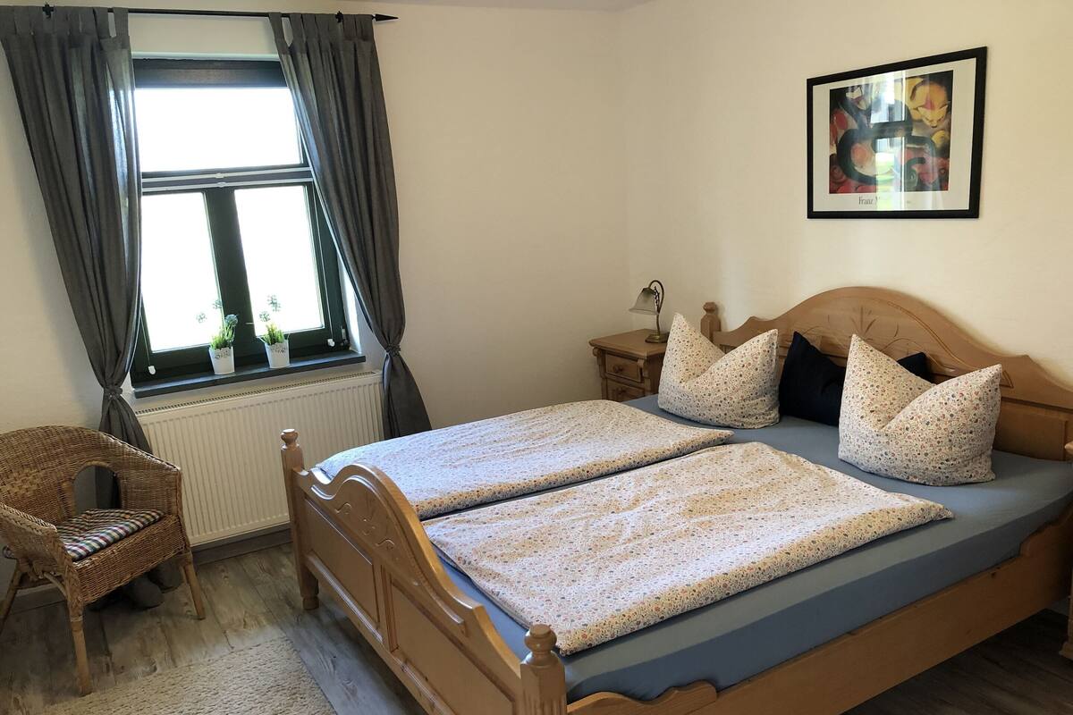Uma cama de casal com roupa de cama floral, uma cabeceira de madeira, uma mesa de cabeceira, uma cadeira de vime, uma janela com cortinas e um quadro na parede.