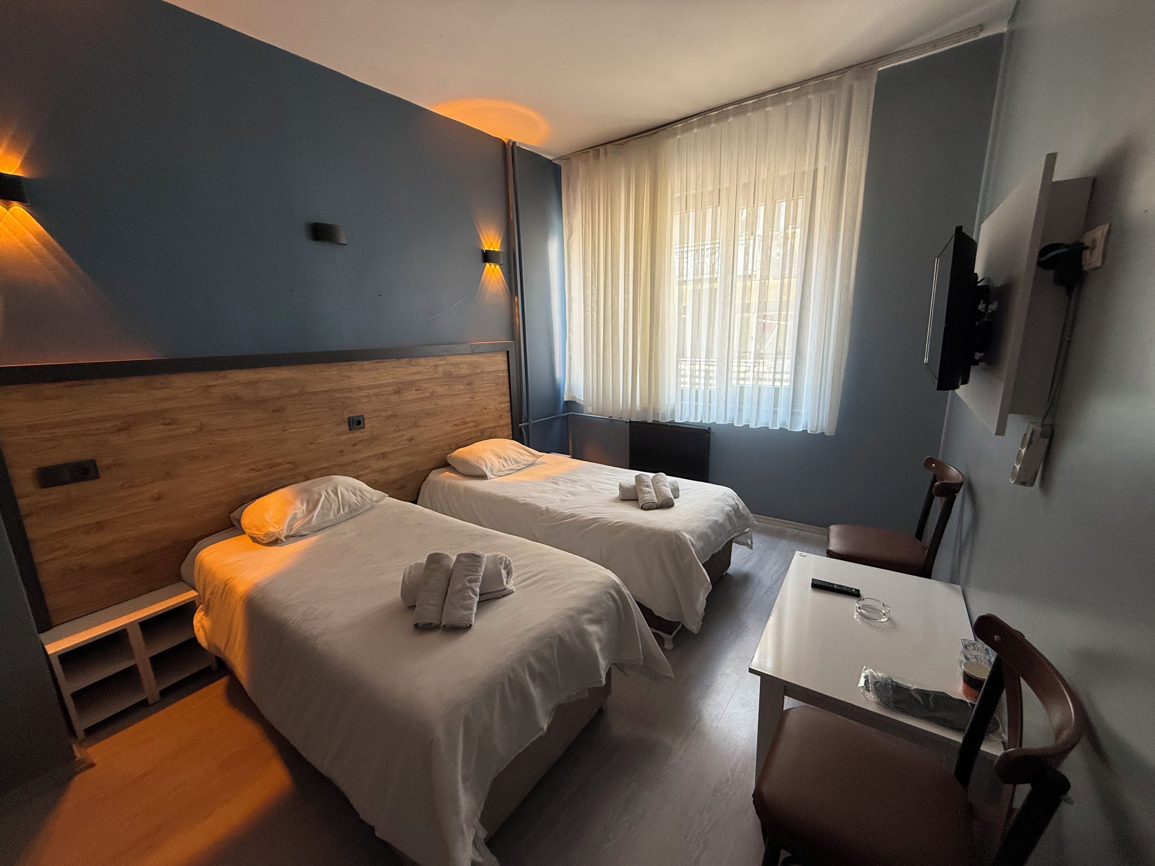 Quarto de hotel com duas camas, uma mesa pequena, uma cadeira, televisão e janela com cortinas.