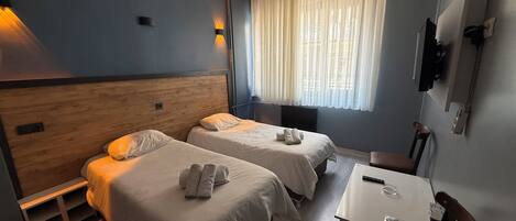 Quarto de hotel com duas camas, uma mesa pequena, uma cadeira, televisão e janela com cortinas.