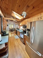 Interior de uma cabana de madeira com cozinha equipada com eletrodomésticos de aço inoxidável, área de jantar com mesa e cadeiras, e uma janela grande que permite entrada de luz natural.