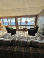 Sala de estar com uma grande janela que oferece vista para o mar. Há duas poltronas pretas e um sofá com estampa xadrez. Uma mesa de centro de madeira está no centro.