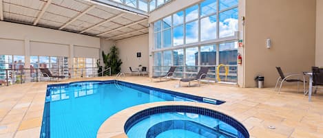 Piscina interna com teto de vidro, espreguiçadeiras e vista da cidade.
