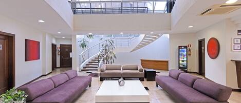 Um hall amplo com teto alto, área de estar central com sofás, balcão de recepção e escadaria para um andar superior.