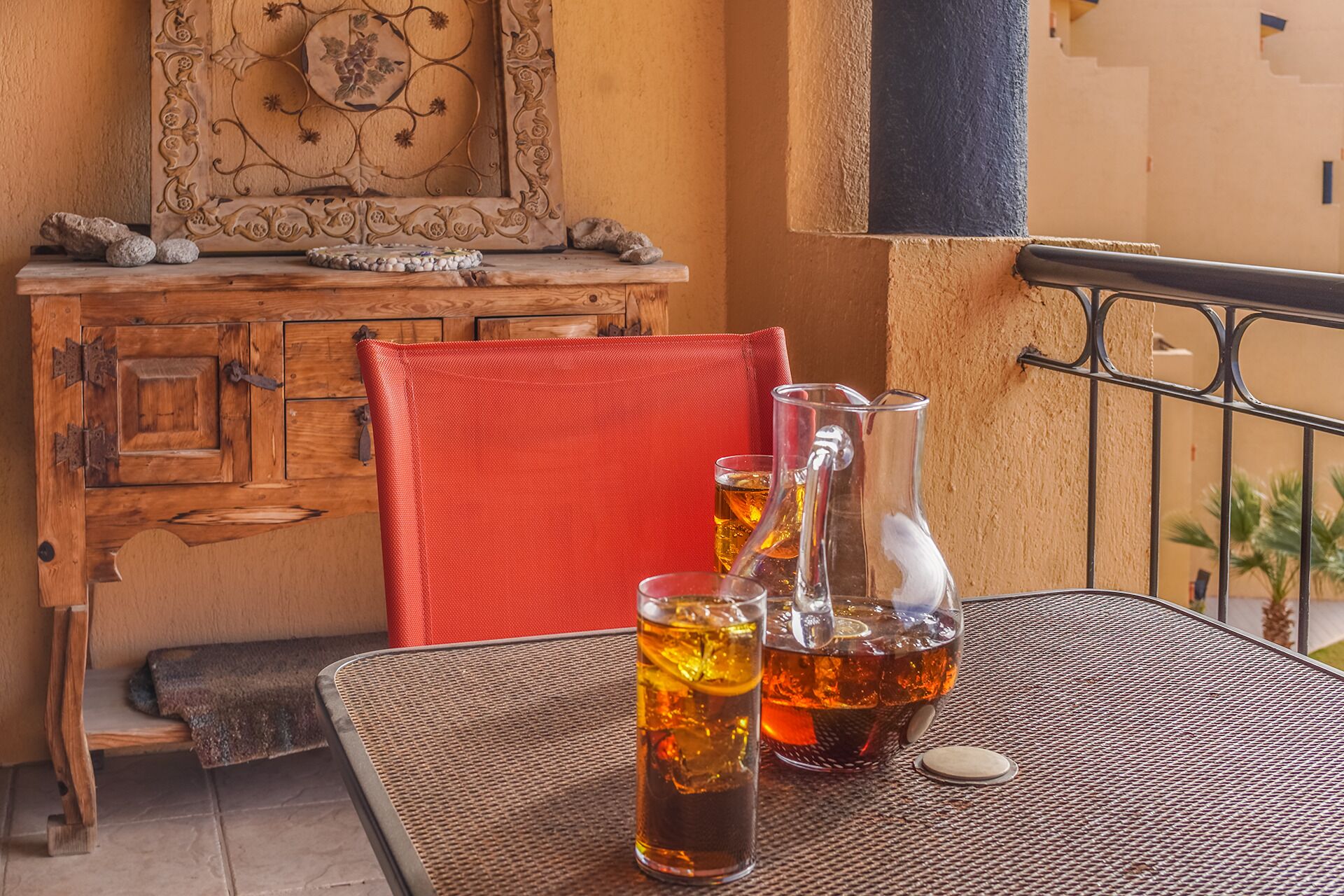 Uma mesa com toalha de tecido trançado, dois copos com líquido âmbar e um jarro de vidro transparente numa varanda, com um armário de madeira e uma cadeira vermelha ao fundo.