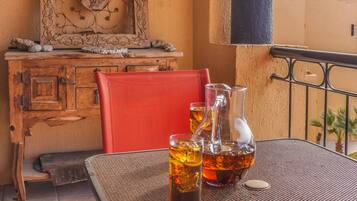 Uma mesa com toalha de tecido trançado, dois copos com líquido âmbar e um jarro de vidro transparente numa varanda, com um armário de madeira e uma cadeira vermelha ao fundo.