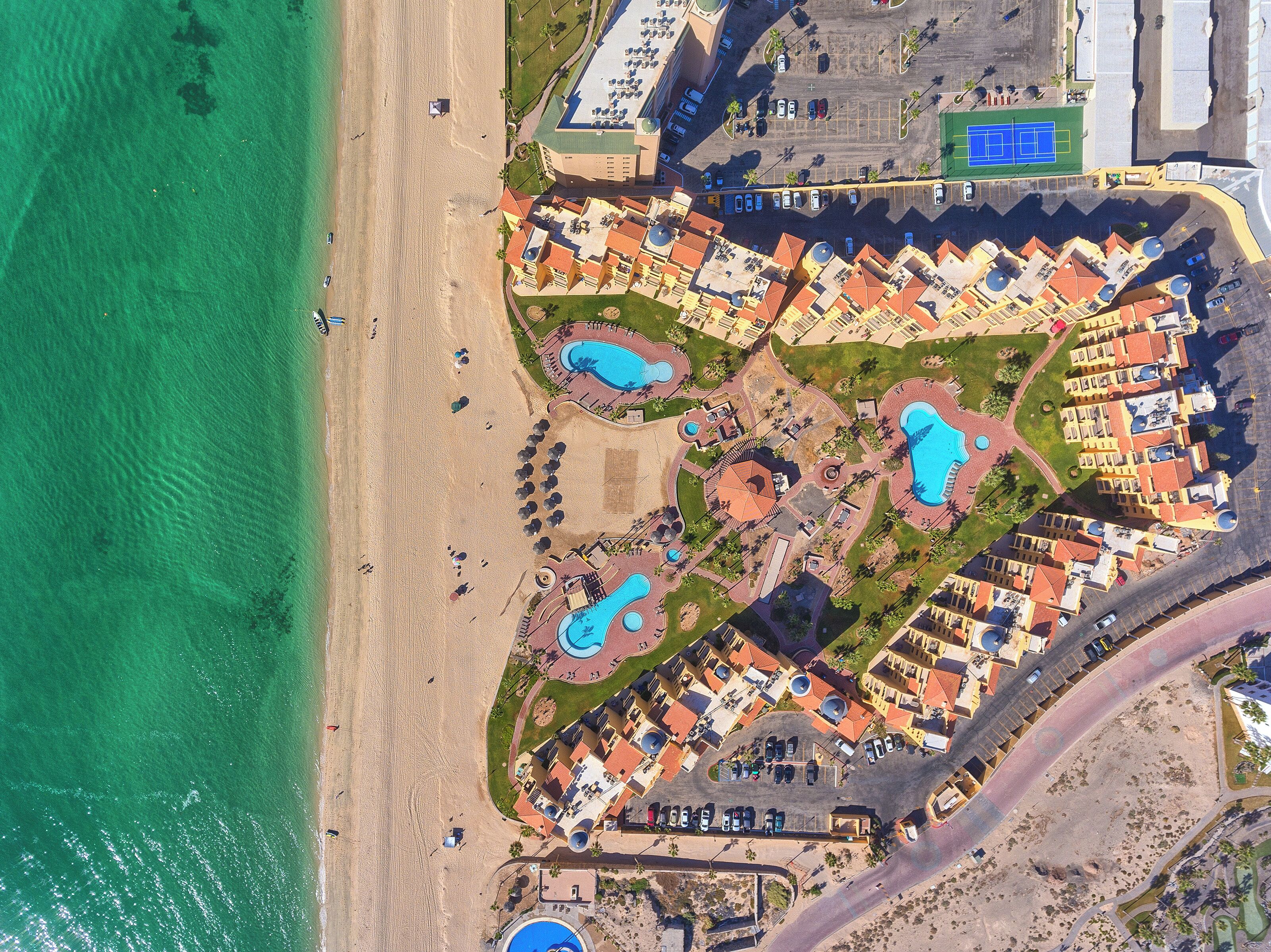 Vista aérea de um resort com várias piscinas, uma praia e estacionamento.