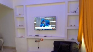 Uma sala de estar com uma TV de tela plana na parede, um sofá de cor escura, uma pequena mesa de centro e prateleiras embutidas com diversos objetos.