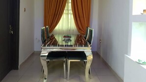 Uma sala de jantar com mesa de vidro e cadeiras, cortinas laranja e uma porta à esquerda.