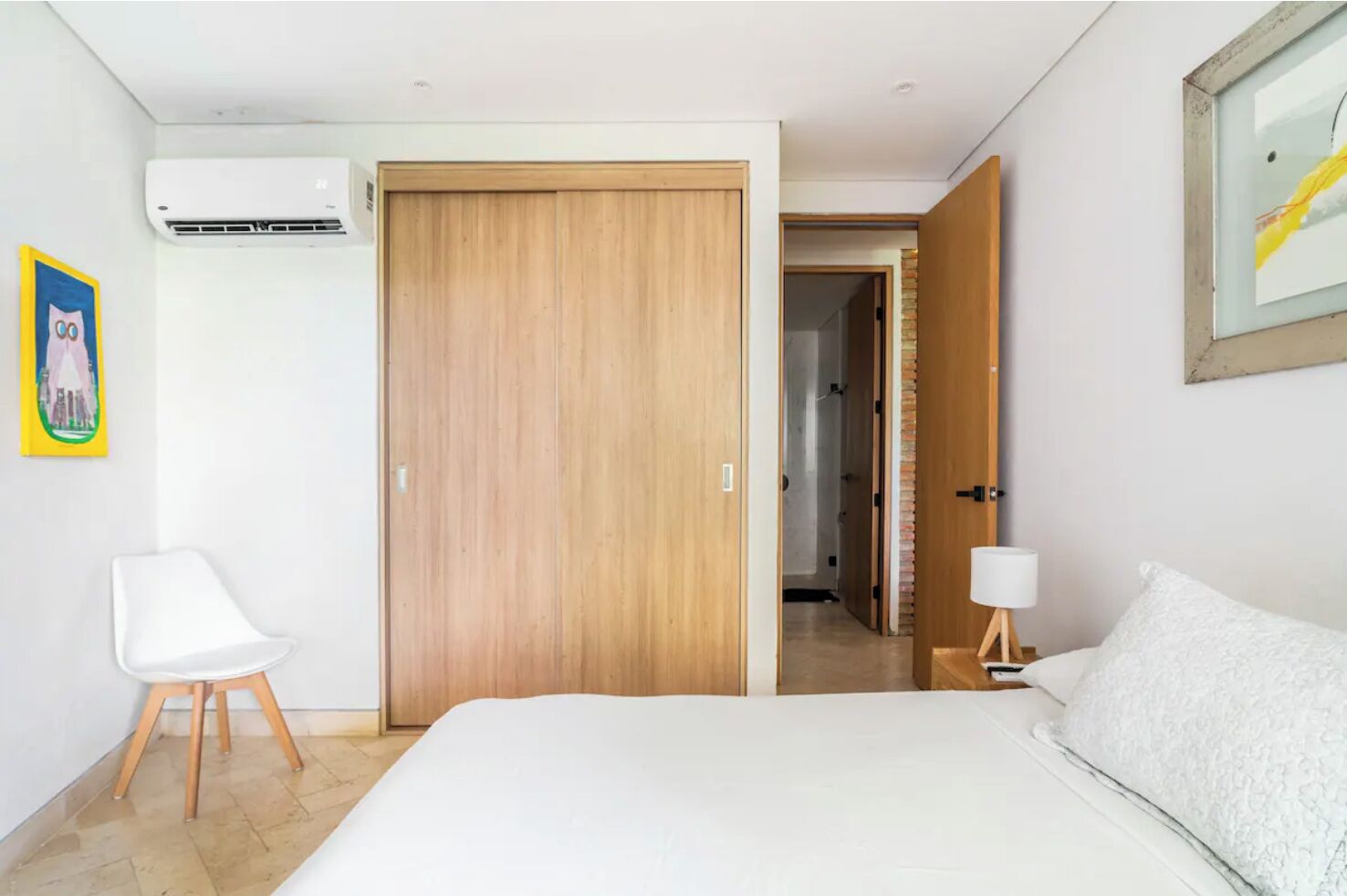 Quarto com porta de madeira, uma cadeira branca, mesa de cabeceira com abajur e uma cama.