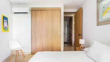 Quarto com porta de madeira, uma cadeira branca, mesa de cabeceira com abajur e uma cama.