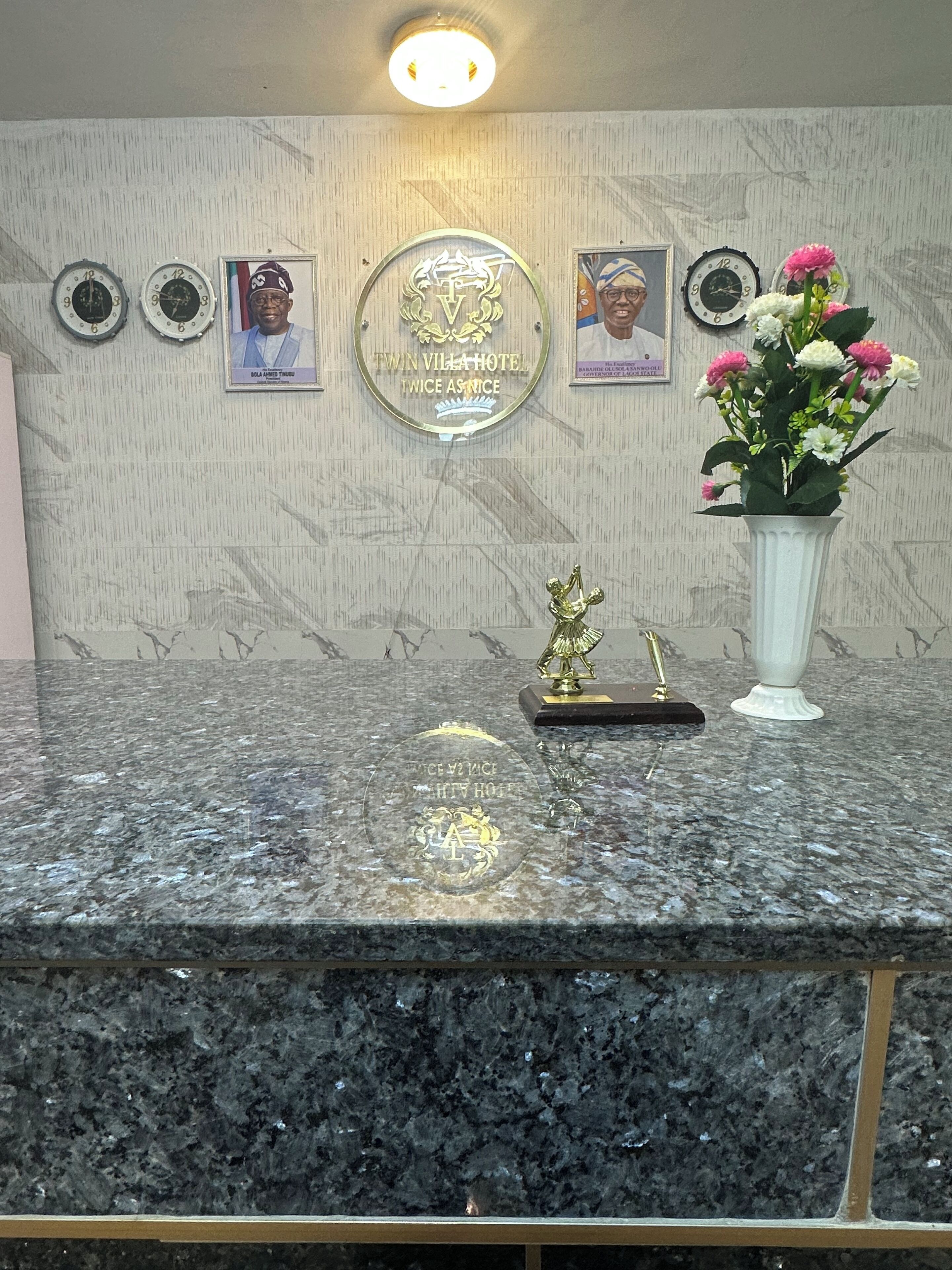 Uma mesa de recepção com tampo de mármore, uma placa com o nome do hotel, fotografias emolduradas e um vaso com flores.
