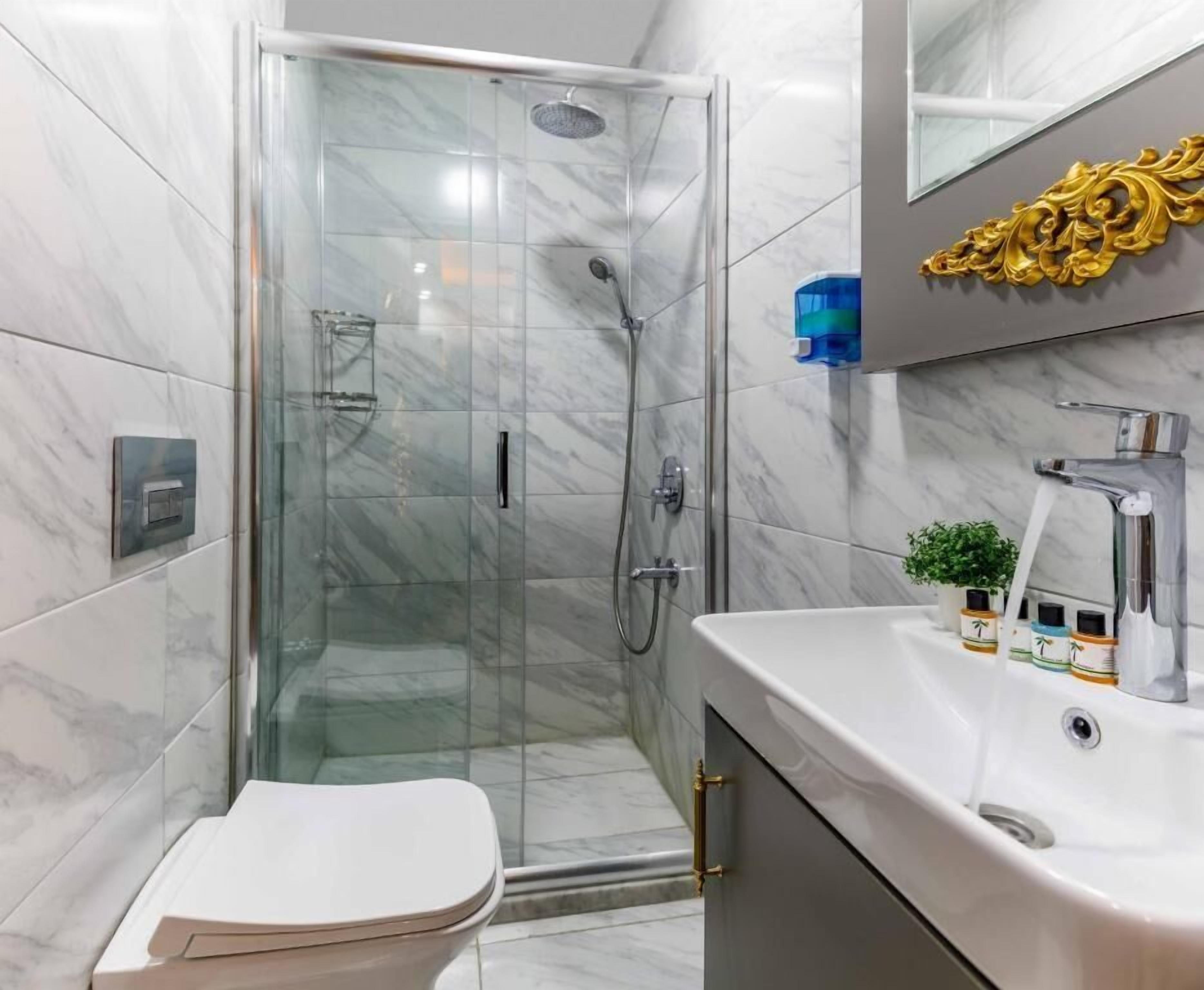 Uma casa de banho moderna com chuveiro em cabine de vidro, sanita branca e lavatório com um detalhe dourado.