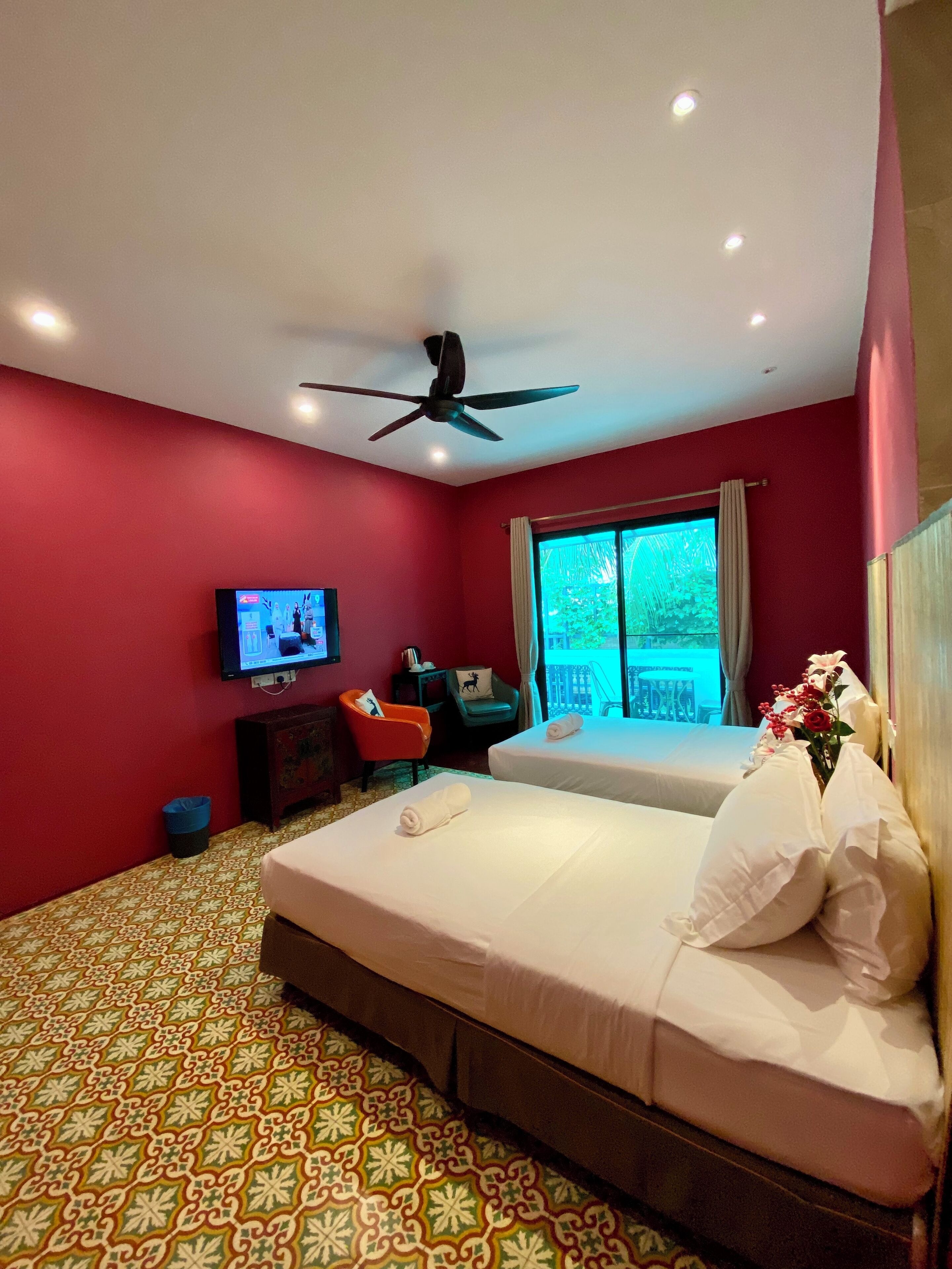 Quarto de hotel com duas camas, televisão, ventoinha no teto e janela com cortinas.