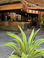 Uma área de bar ao ar livre com balcão de madeira, assentos e diversas plantas.