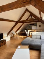 Um quarto de sótão aconchegante, com vigas de madeira aparentes, um sofá cinza, uma mesa de jantar de madeira e uma televisão.