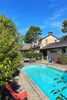 Uma piscina com espreguiçadeiras, uma casa com telhado inclinado e um céu azul límpido.