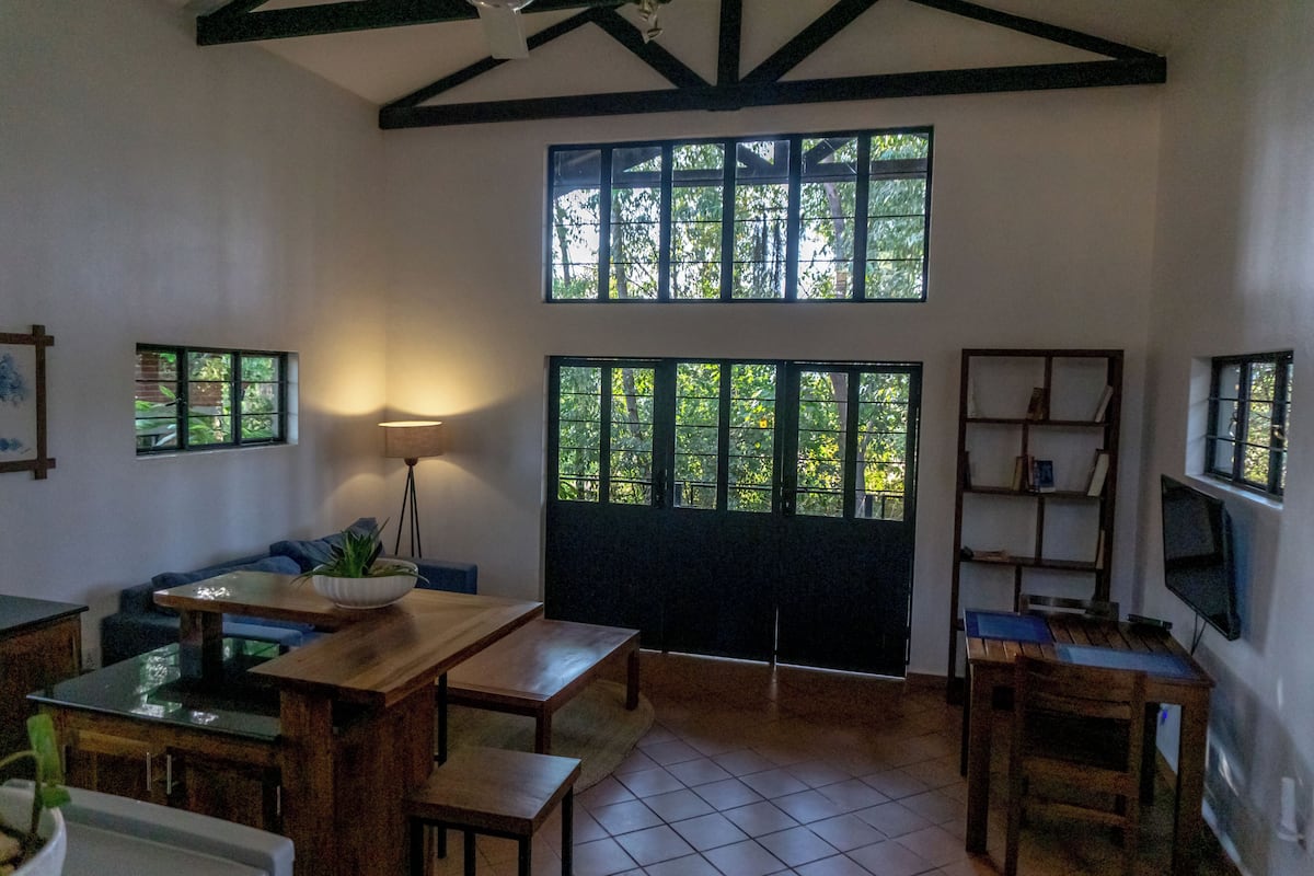 Uma sala de estar ampla com teto alto, grandes janelas, uma mesa de jantar de madeira, um sofá e uma TV fixada na parede.
