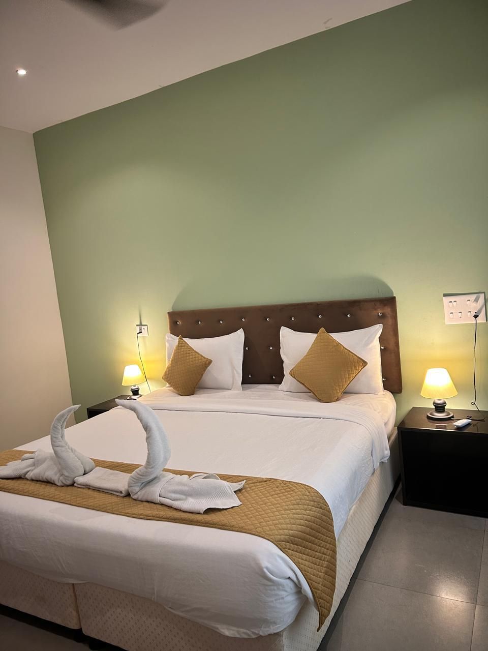 Uma cama bem arrumada com cabeceira marrom, duas luminárias de apoio e um par de toalhas em formato de cisne sobre a cama.
