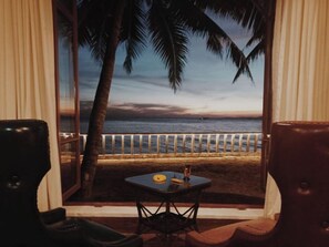 Quarto com vista para a praia e oceano ao pôr do sol, com uma pequena mesa, uma garrafa e um prato.