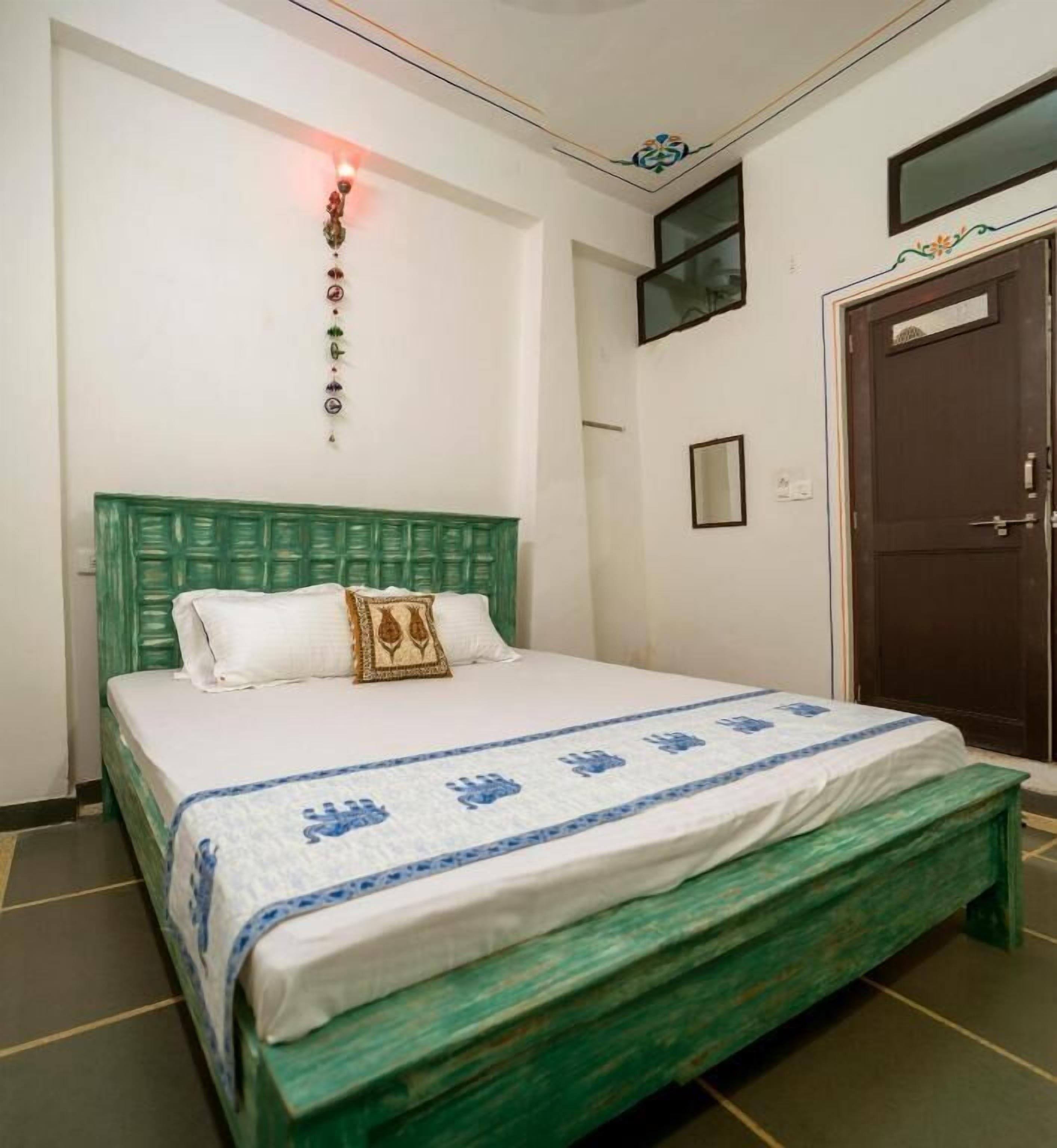 Quarto com cabeceira verde, cama com colcha estampada em branco e azul, luminária fixada na parede, um pequeno espelho e uma porta com moldura decorativa.