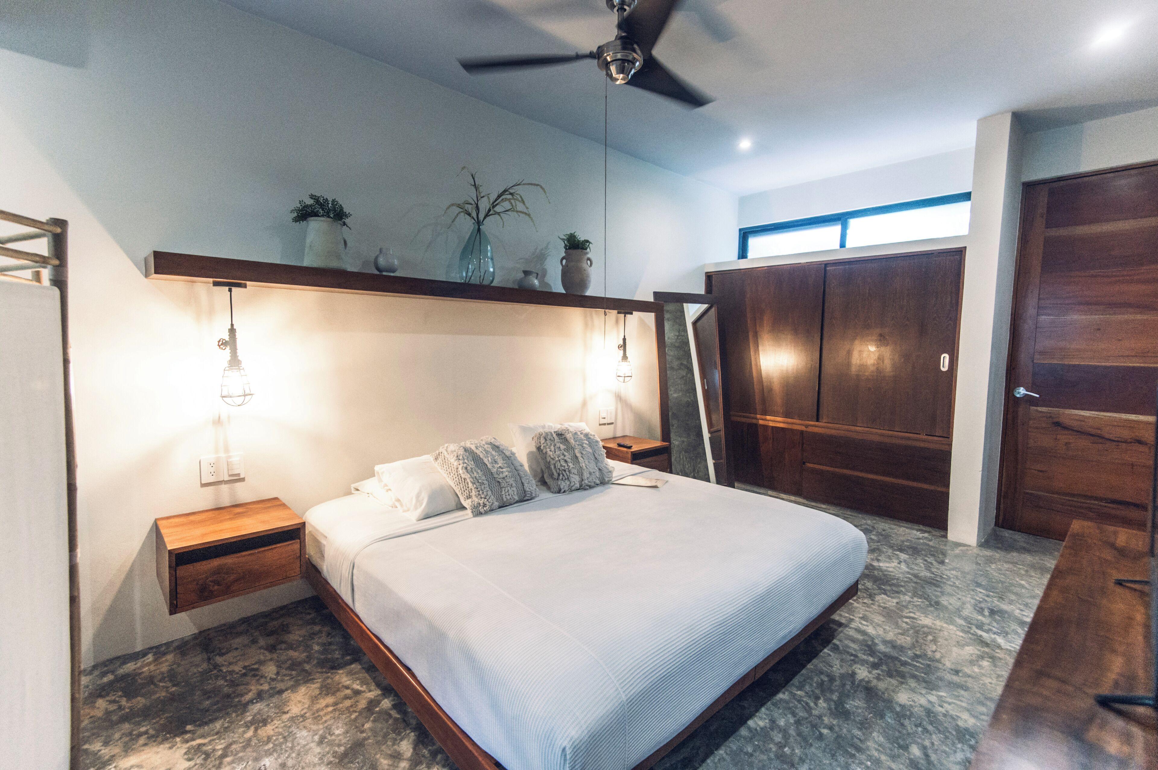 Quarto com cama, luminárias de cabeceira, ventoinha no teto, guarda-roupa de madeira e prateleira com objetos decorativos.