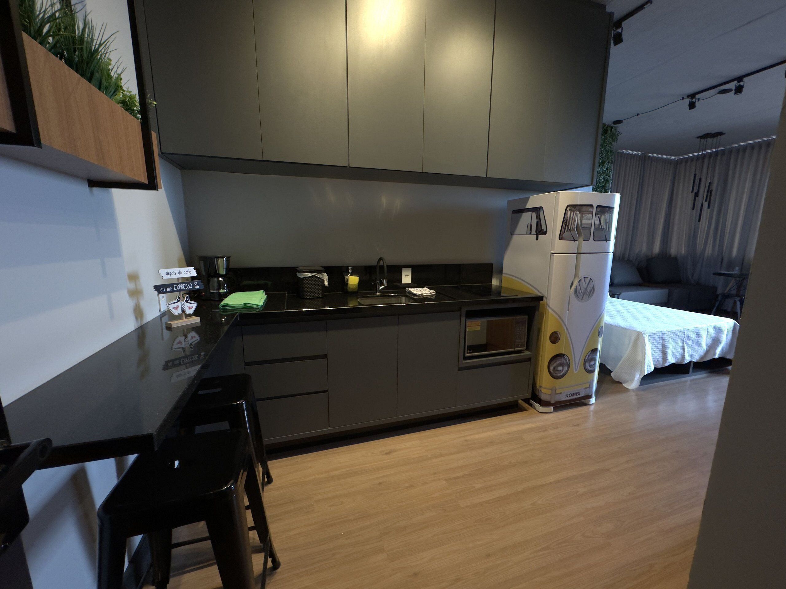 Cozinha compacta com bancada preta, pia pequena e geladeira compacta. Uma cama com cobertura branca aparece ao fundo.