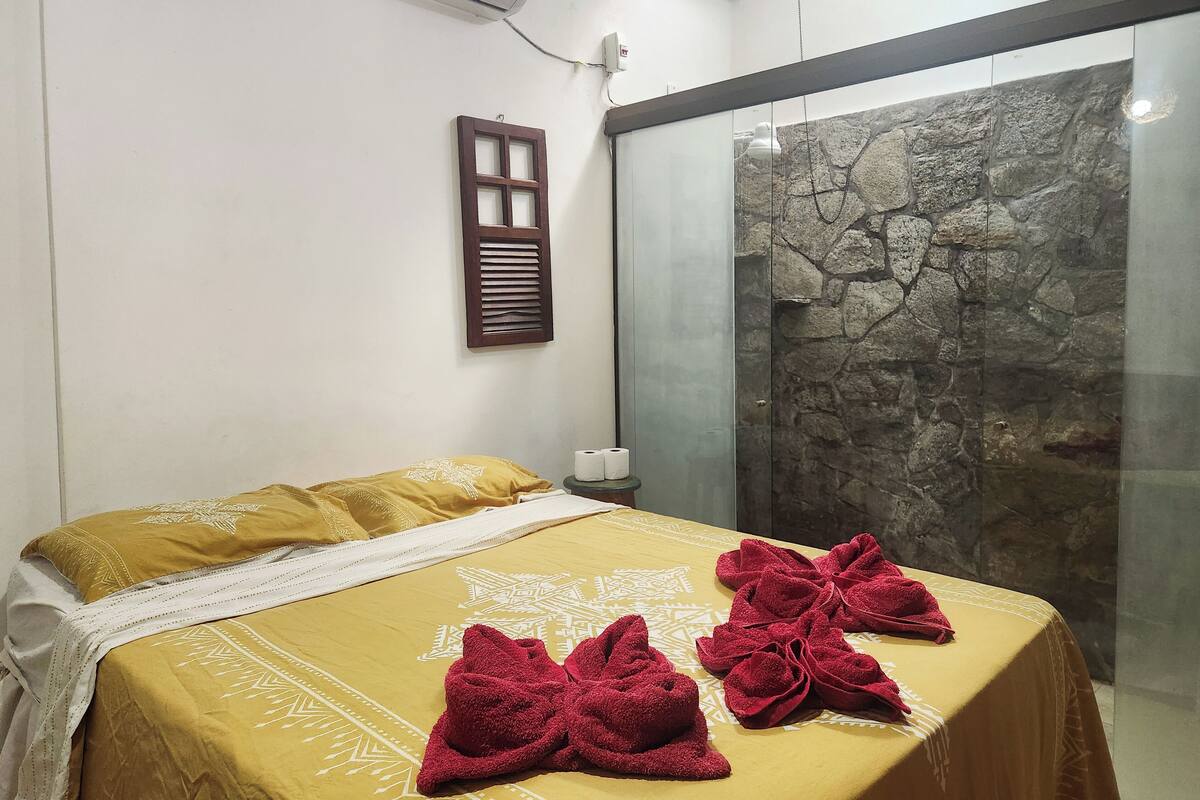 Quarto de hotel com uma cama, janela com parede de pedra, ar condicionado e uma pequena mesa com toalhas.