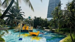 Piscina com escorregador amarelo e estrutura de recreação, cercada por palmeiras e vegetação.