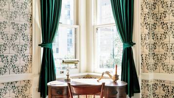 Quarto de canto com papel de parede estampado, uma escrivaninha de madeira com gavetas, uma cadeira, uma janela com cortinas verdes e um objeto decorativo no parapeito.