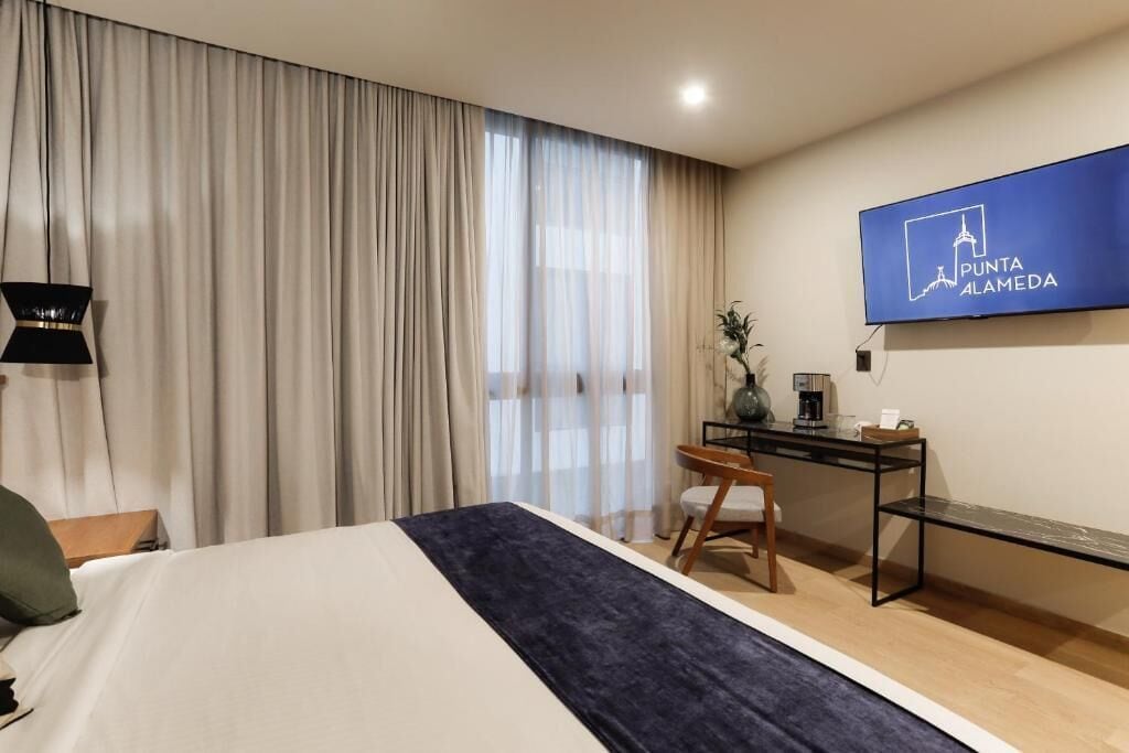Quarto de hotel com cama, escrivaninha, cadeira, televisão e janela com cortinas.