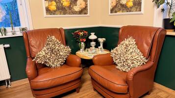 Duas poltronas de couro marrom com serragem como almofadas, uma mesa redonda pequena com um vaso de flores, e dois quadros florais emoldurados na parede.