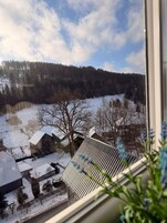 Vista da janela sobre uma paisagem nevada com casas, floresta e céu claro.